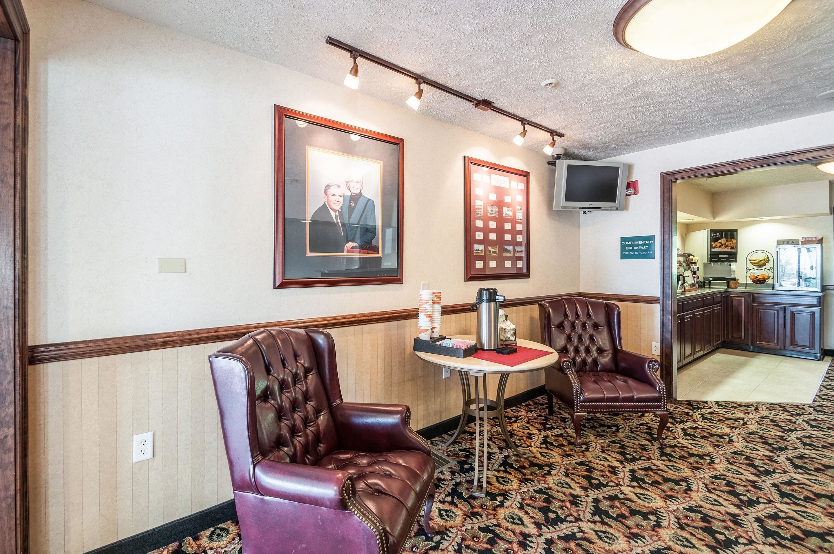 Um hall de hotel com duas poltronas de couro, uma mesa com uma cafeteira, quadros nas paredes e uma cozinha visível através de uma porta aberta.