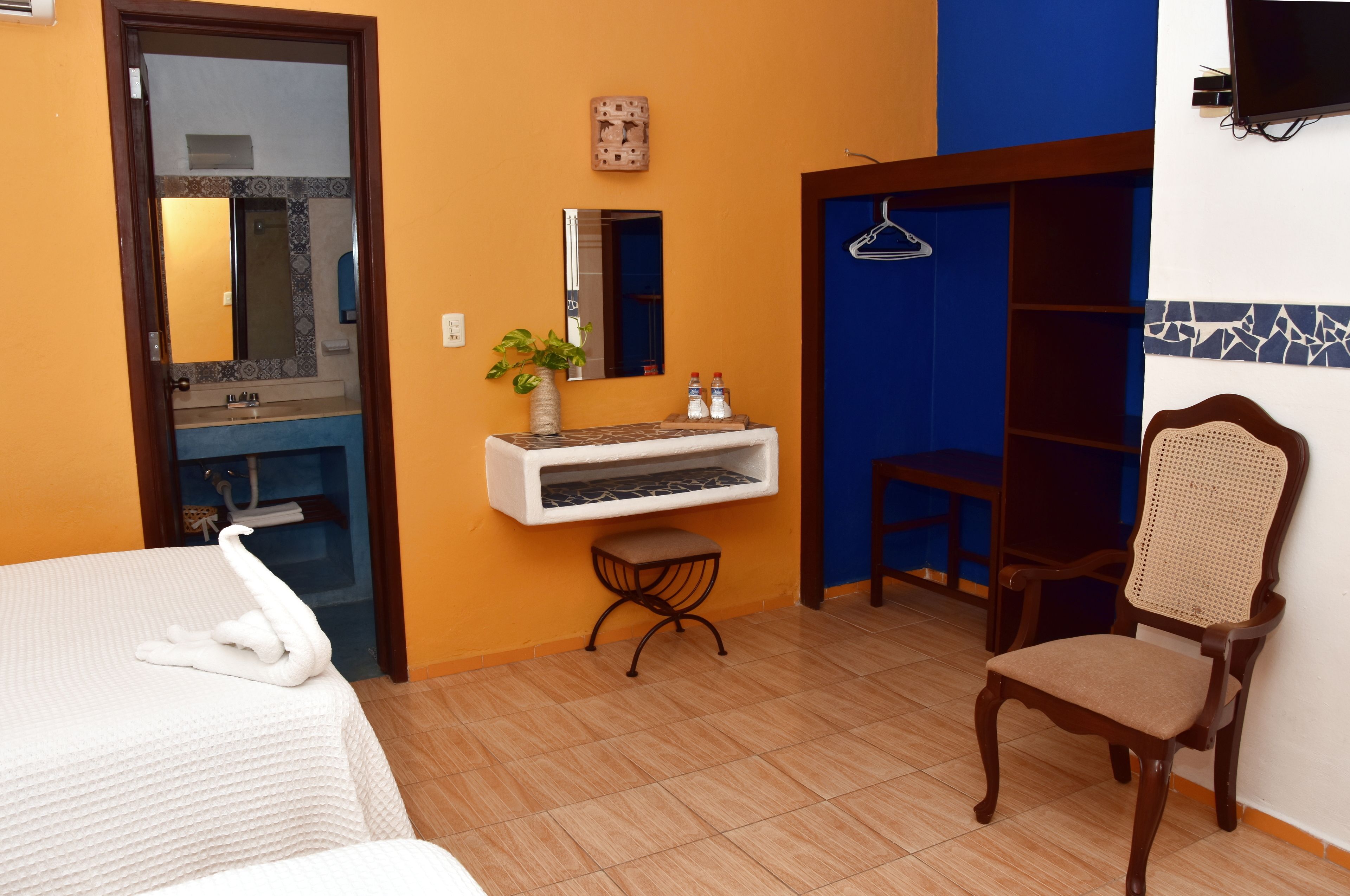 Quarto de hotel com uma cama, uma toucador com espelho, uma pequena mesa com planta, uma cadeira e um guarda-roupa.