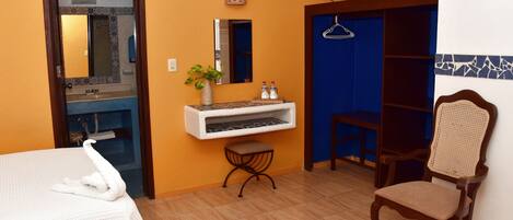 Quarto de hotel com uma cama, uma toucador com espelho, uma pequena mesa com planta, uma cadeira e um guarda-roupa.
