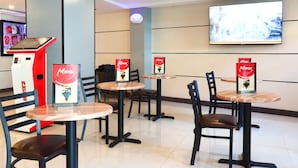 Interior de restaurante com mesas, cadeiras, suporte para cardápio, tela de televisão e máquina de vendas automática.