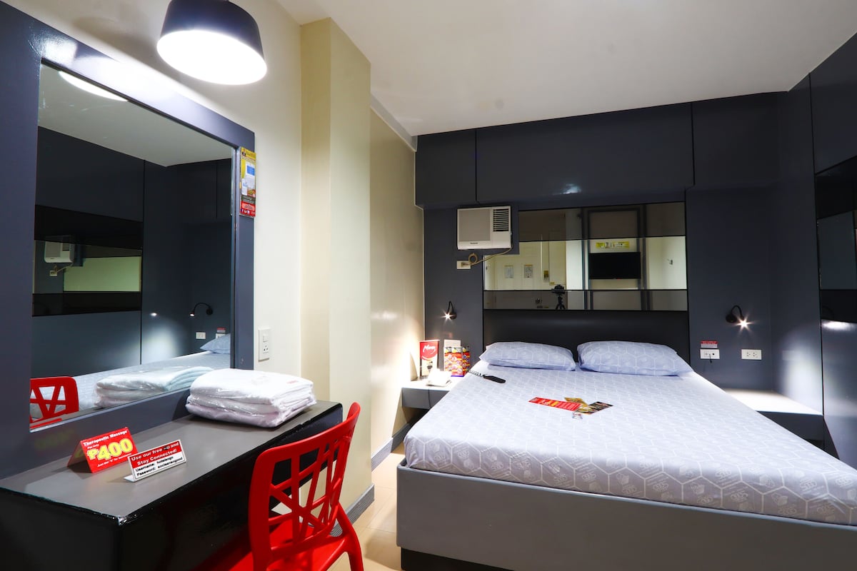 Quarto de hotel com cama, escrivaninha, cadeira, espelho grande e mesa de cabeceira.