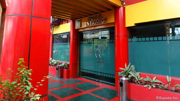 Entrada de hotel com fachada em vermelho e amarelo, portas de vidro e piso de azulejos.