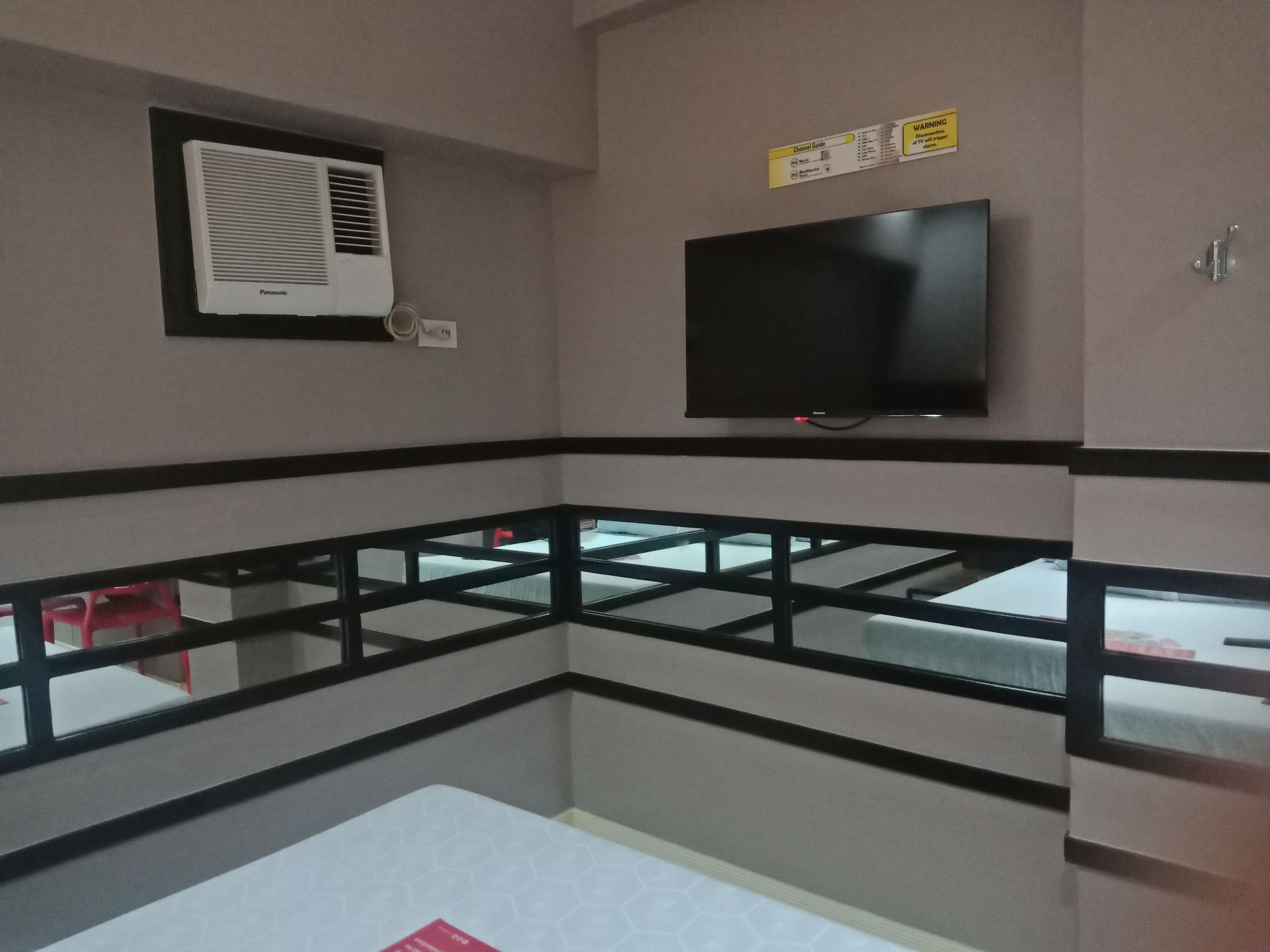 Quarto de hotel com televisão de tela plana na parede, ar-condicionado Panasonic e divisória de vidro com moldura preta.
