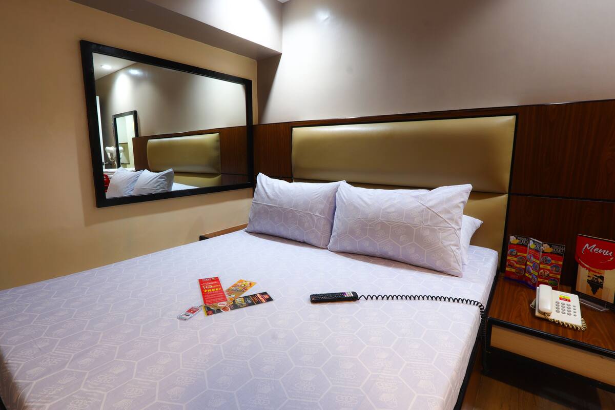 Quarto de hotel com uma cama grande, uma mesa de cabeceira com telefone e um sinal de menu, um espelho e uma parede com padrão geométrico.