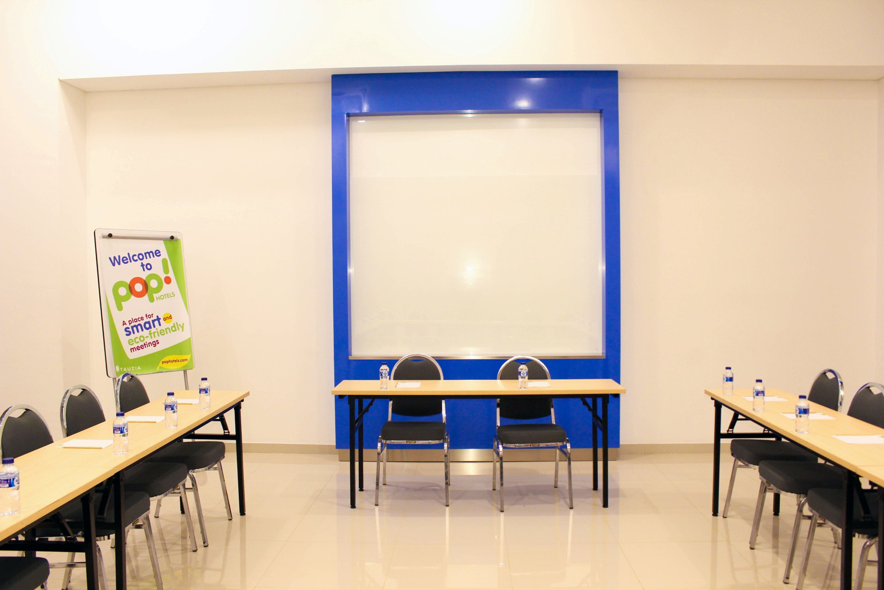 Uma sala de conferências com um quadro branco grande, duas mesas com cadeiras e um aviso promovendo reuniões ecologicamente responsáveis.