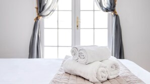 Cama bem arrumada com toalhas brancas dobradas, janela com cortinas e uma porta ao fundo.