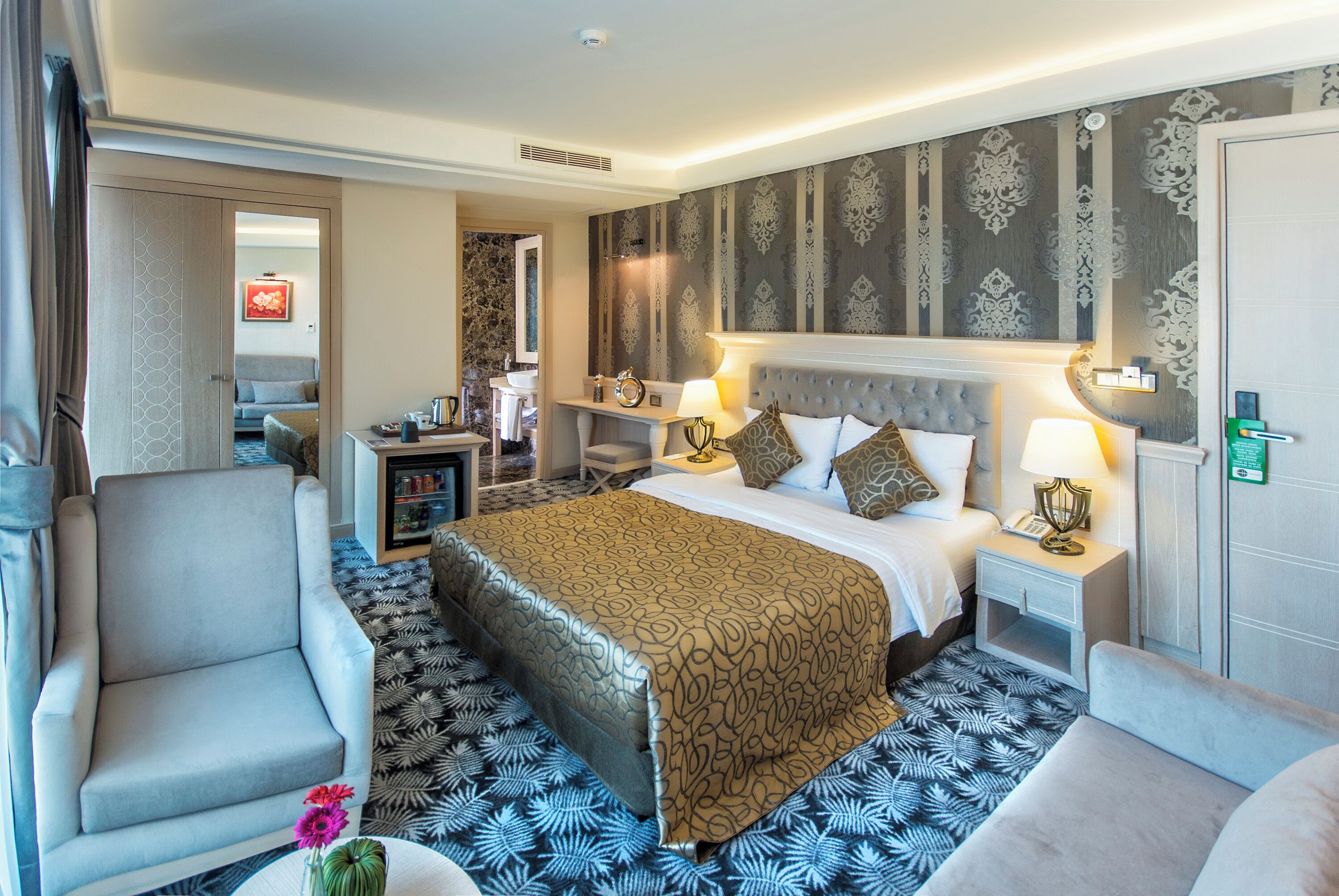 Quarto de hotel com uma cama grande, área de estar com uma poltrona, mesa de cabeceira com abajur e um guarda-roupa.