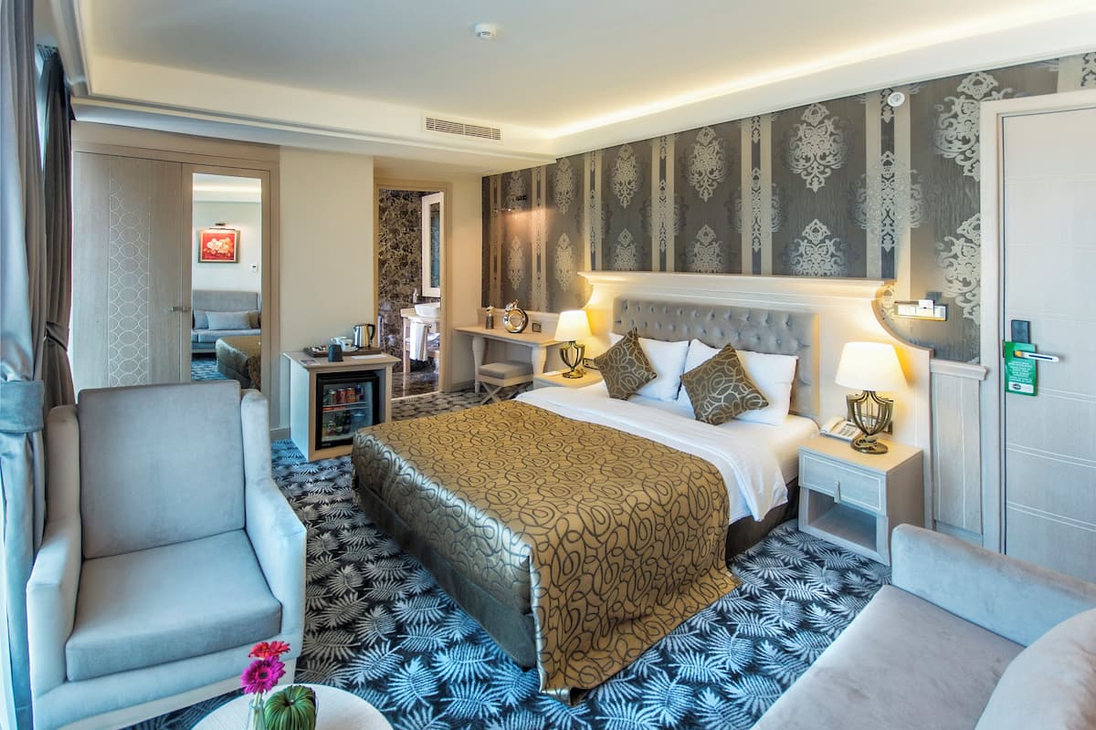 Quarto de hotel com uma cama grande, área de estar com uma poltrona, mesa de cabeceira com abajur e um guarda-roupa.