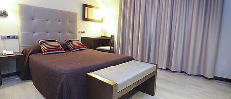 Quarto de hotel moderno com uma cama grande, mesa de cabeceira, escrivaninha e uma janela ampla com cortinas.