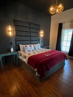 Quarto com cama grande, cabeceira escura, luminárias de cabeceira, mesa de apoio e janela com cortinas.