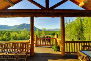 Um deck de madeira com cadeiras e guarda-corpo com vista para uma floresta e montanhas.