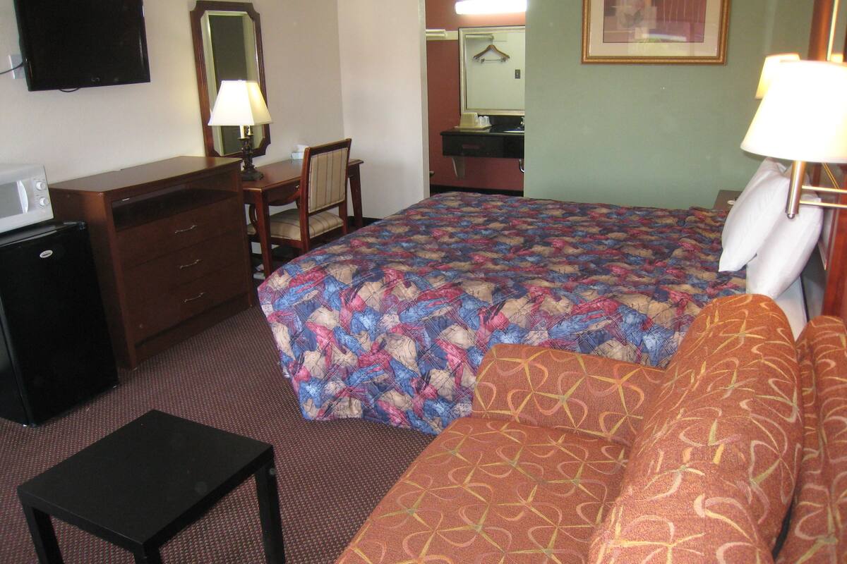 Quarto de hotel com uma cama, escrivaninha, cadeira, mesinha, televisão, espelho e abajur.