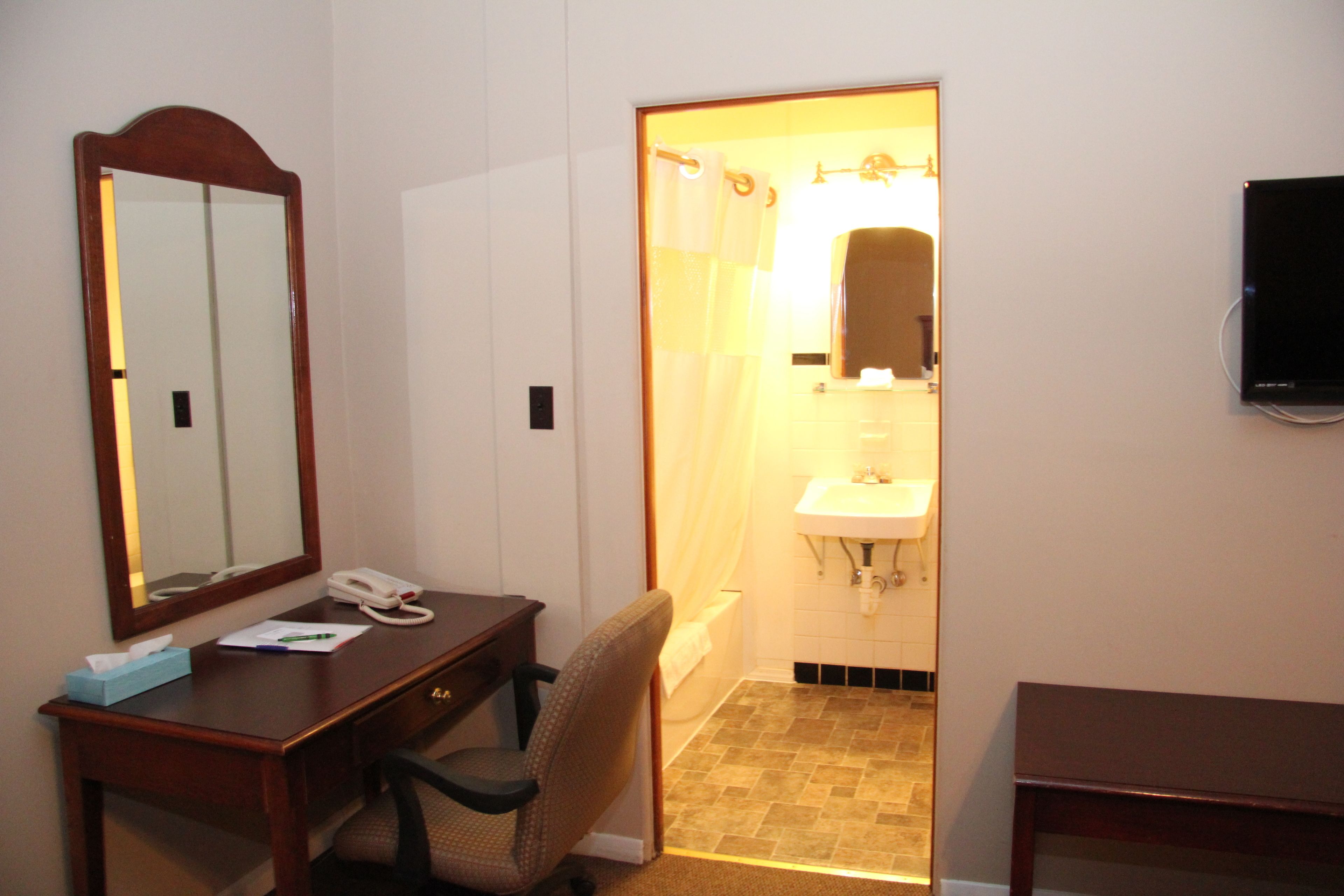Quarto de hotel com escrivaninha, cadeira, espelho e banheiro visível por uma porta aberta.