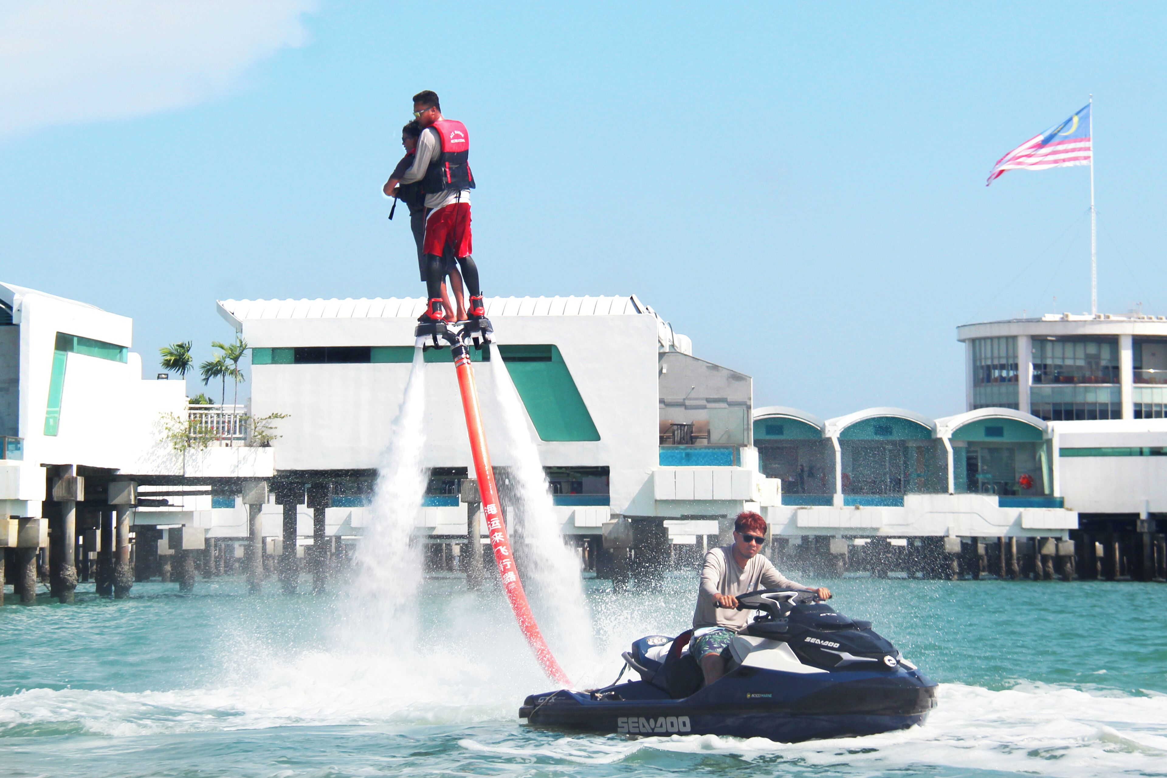 Um jet ski com uma pessoa em pé e outra sentada, e um edifício com uma bandeira ao fundo.