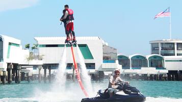 Um jet ski com uma pessoa em pé e outra sentada, e um edifício com uma bandeira ao fundo.