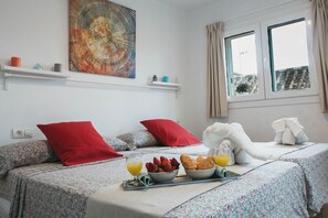 Quarto arrumado com duas camas, bandeja de café da manhã com frutas e suco, e janela com cortinas.
