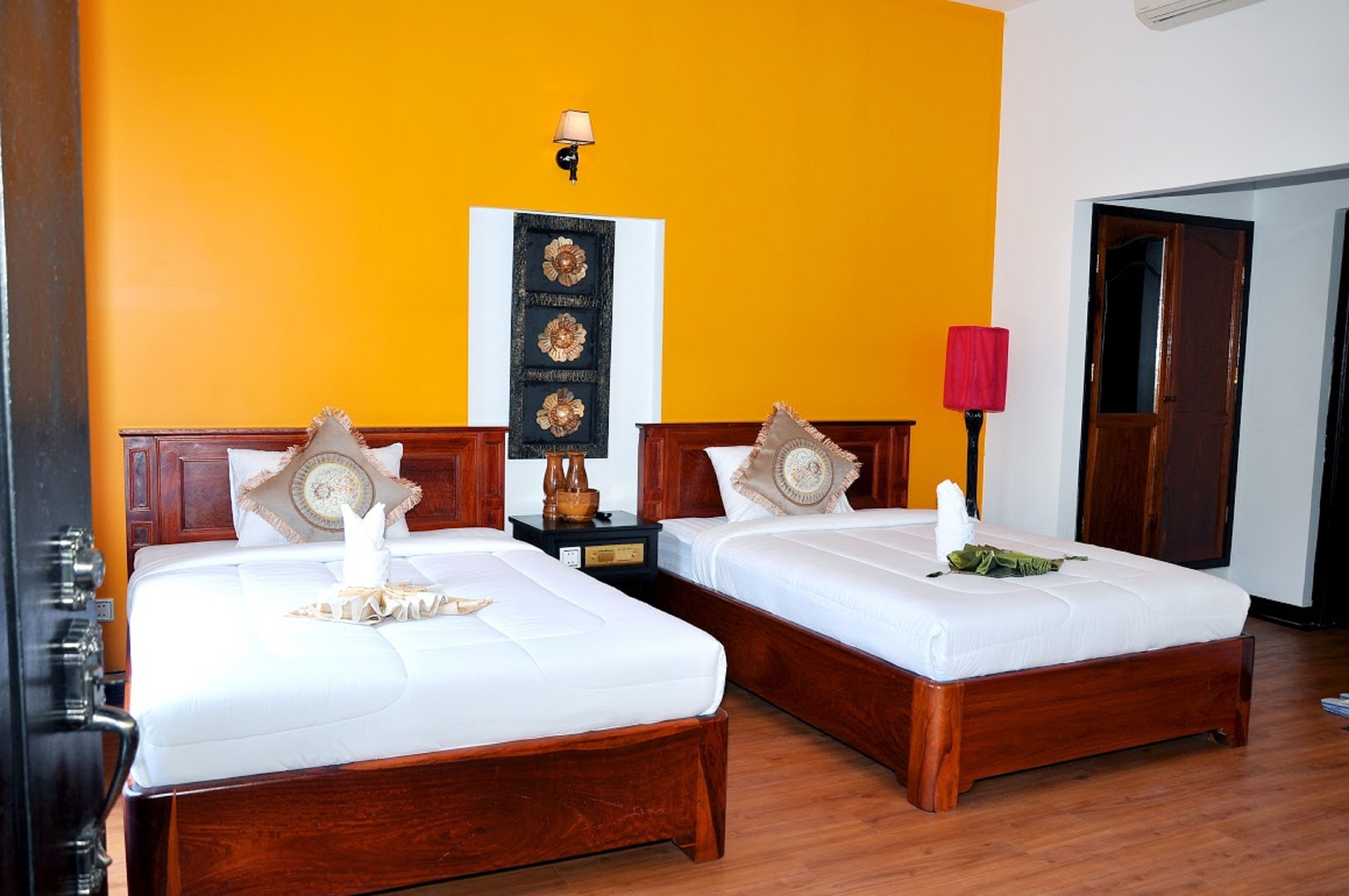 Quarto de hotel com duas camas de madeira, uma mesa de cabeceira com abajur e um painel decorativo fixado na parede.