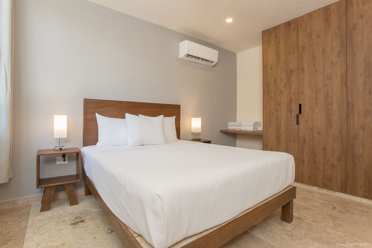 Cama bem arrumada com cabeceira de madeira, duas mesas de cabeceira com luminárias, guarda-roupa de madeira e um aparelho de ar condicionado na parede.
