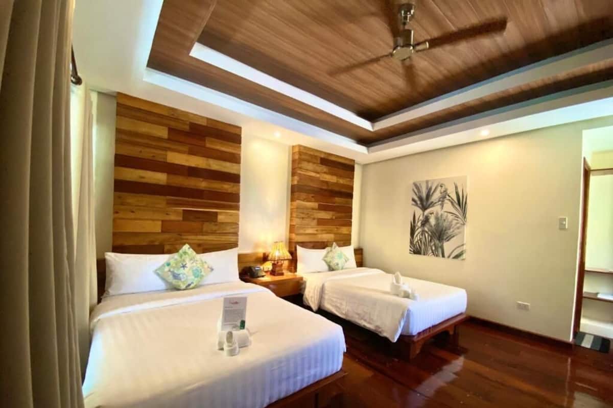 Quarto de hotel com duas camas, cabeceiras de madeira, um ventilador de teto e um quadro na parede.