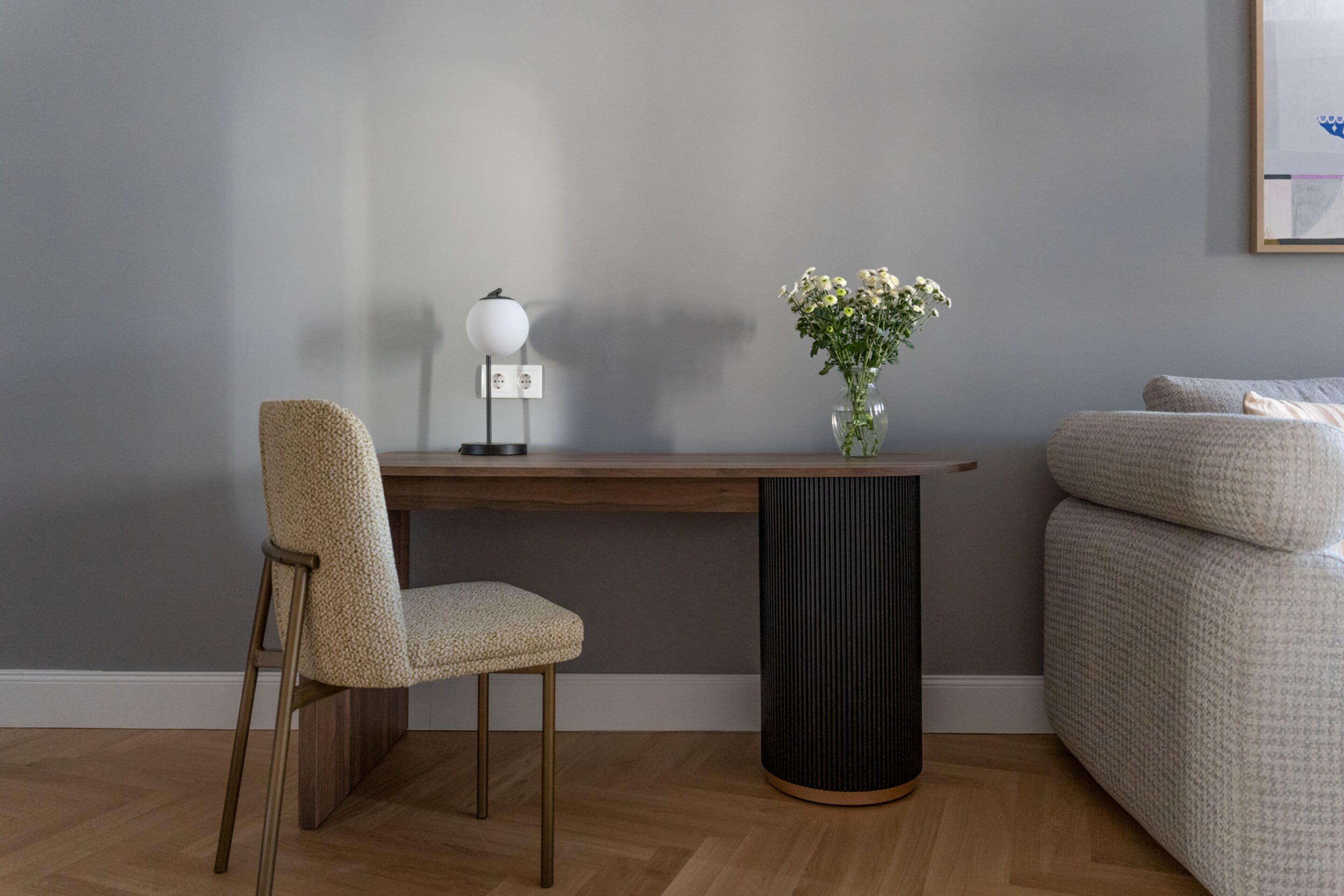 Quarto moderno com escrivaninha de madeira, uma cadeira, uma mesinha com um vaso de flores e um sofá.
