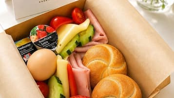 Caixa de pequeno-almoço com um cartão, contendo pão, fruta, queijo e um copo de sumo de laranja.