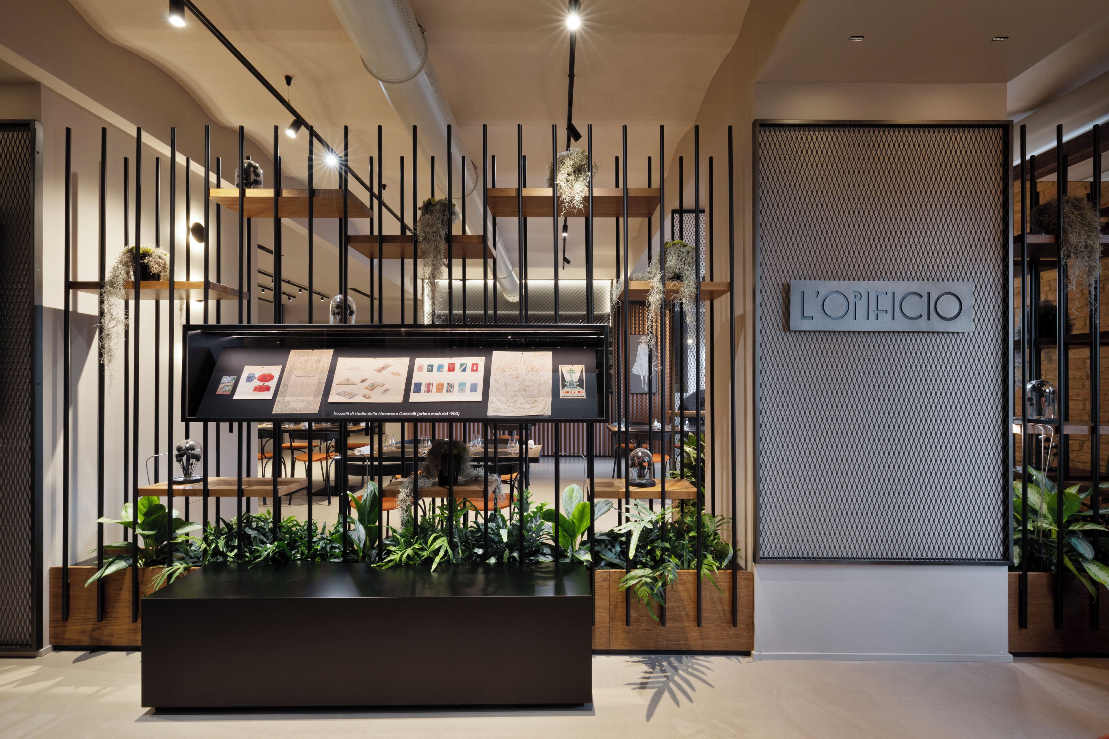 Uma área de recepção com design moderno, apresentando um balcão de atendimento preto, painéis verticais de metal preto e uma placa com a inscrição "L'OPIFICIO".