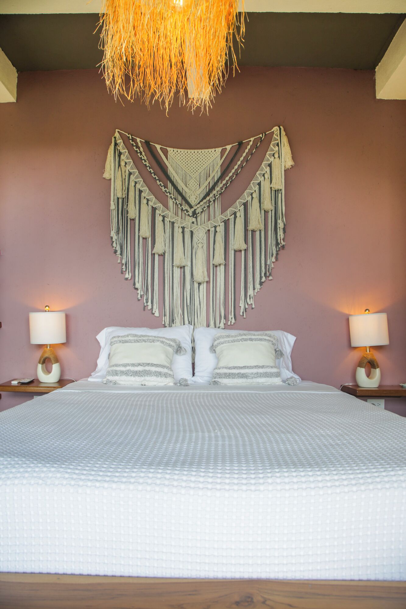 Quarto com uma cama grande, duas mesas de cabeceira com luminárias, um pendente de macramé na parede e um lustre.