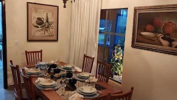 Uma sala de jantar com uma mesa de madeira posta para seis pessoas, rodeada por cadeiras. Há um candelabro sobre a mesa, um quadro emoldurado na parede e uma janela com cortinas.