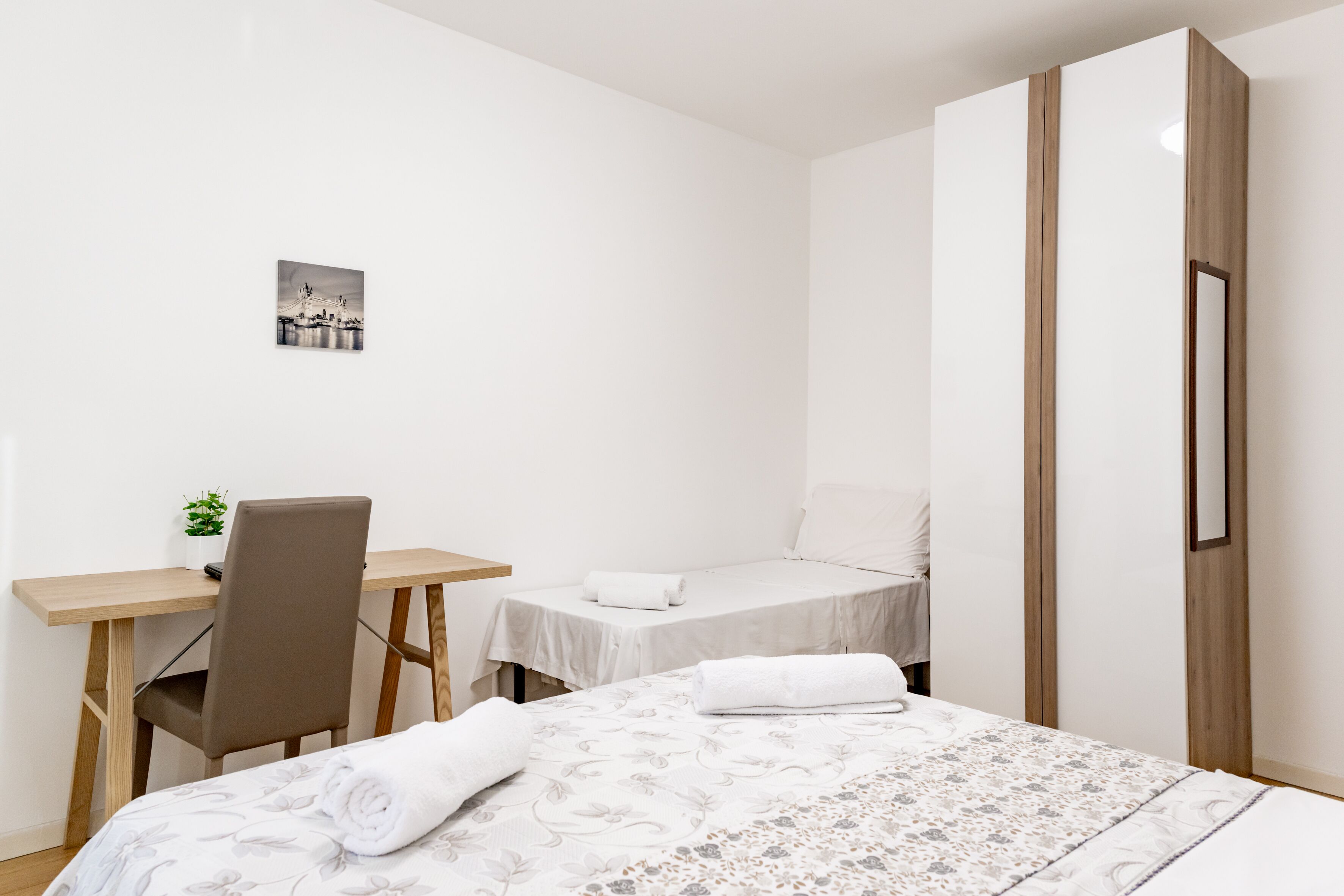 Quarto de hotel com cama, escrivaninha, cadeira, uma pequena planta e um quadro na parede.