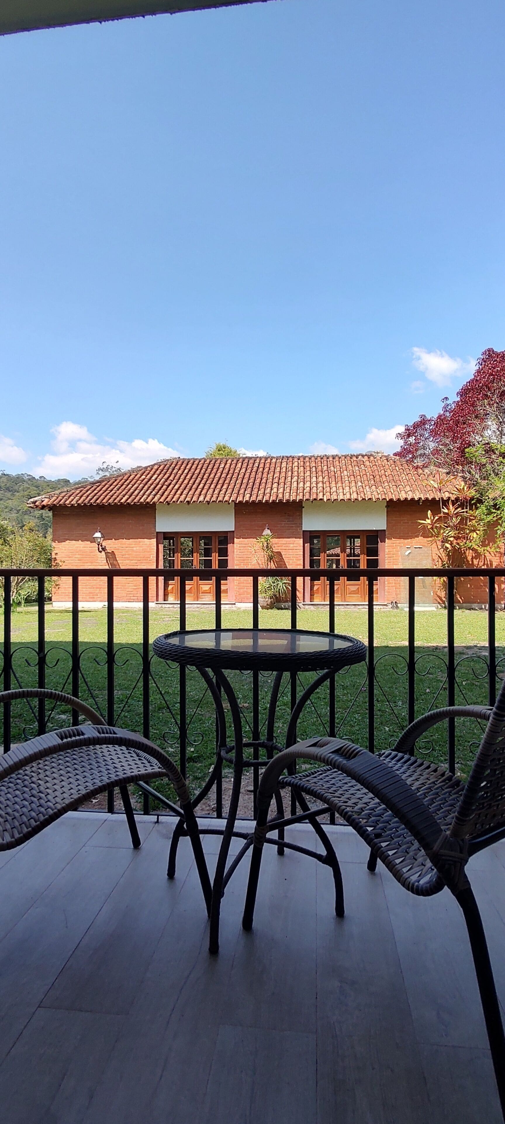 Um pátio com móveis de vime e uma mesa, um prédio de tijolos com telhado de cerâmica ao fundo, e um céu azul límpido.