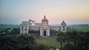 Um edifício grande e histórico com uma cúpula central e alas simétricas, rodeado por palmeiras e campos abertos.