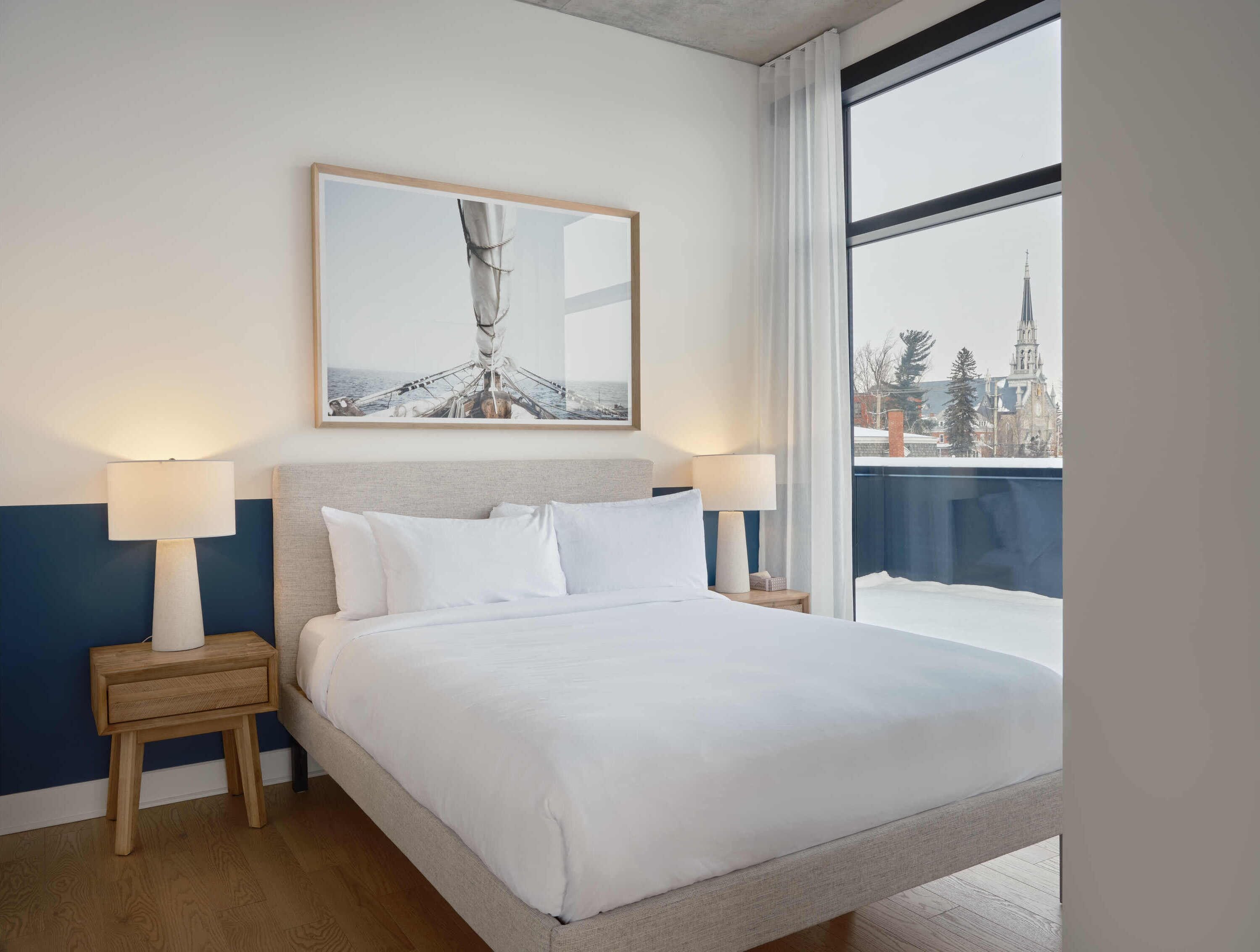 Quarto de hotel com uma cama, duas mesas de cabeceira com abajures, uma janela grande com vista para uma igreja e árvores cobertas de neve.