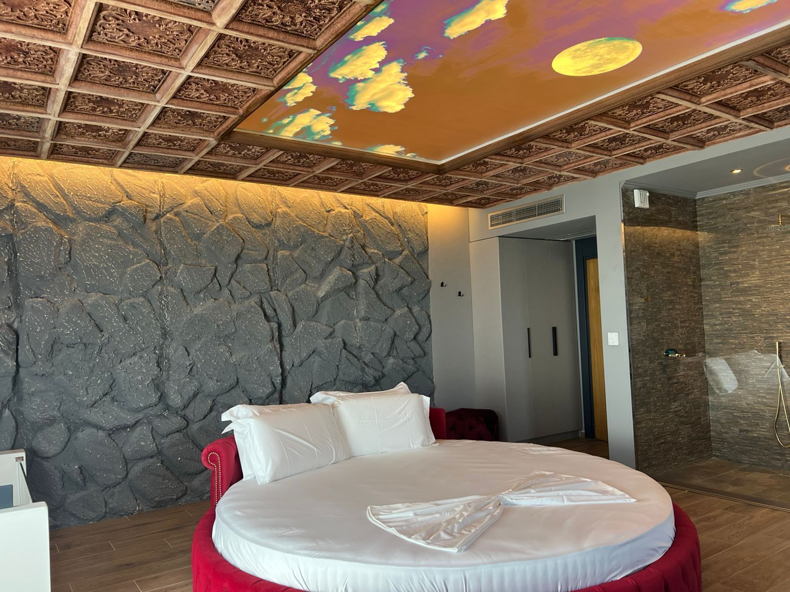 Quarto moderno com parede de pedra, cama redonda com roupa de cama branca e teto com painel decorativo.