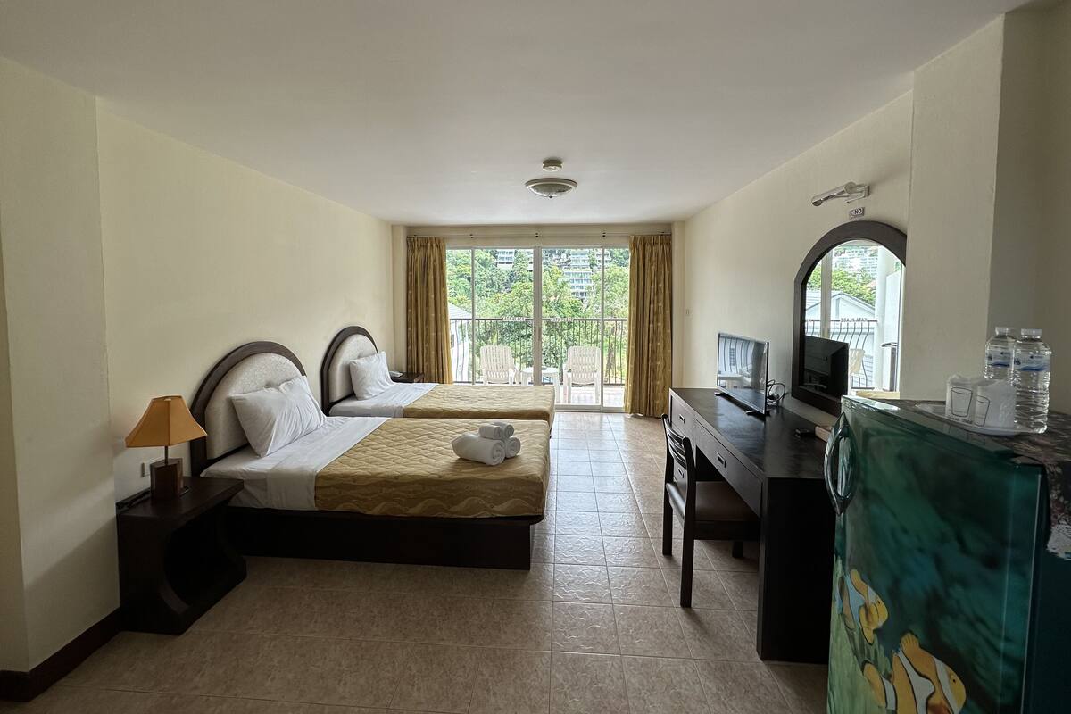 Quarto de hotel com duas camas, uma escrivaninha, televisão e vista para o verde pela janela.