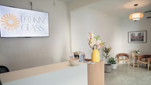 Uma área de recepção com um letreiro 'Looking Glass', um balcão de atendimento, cadeiras, um vaso de planta e uma moldura com uma imagem na parede.