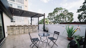Um pátio externo com mesa e cadeiras, pérgola, luzes de corda e plantas em vasos.