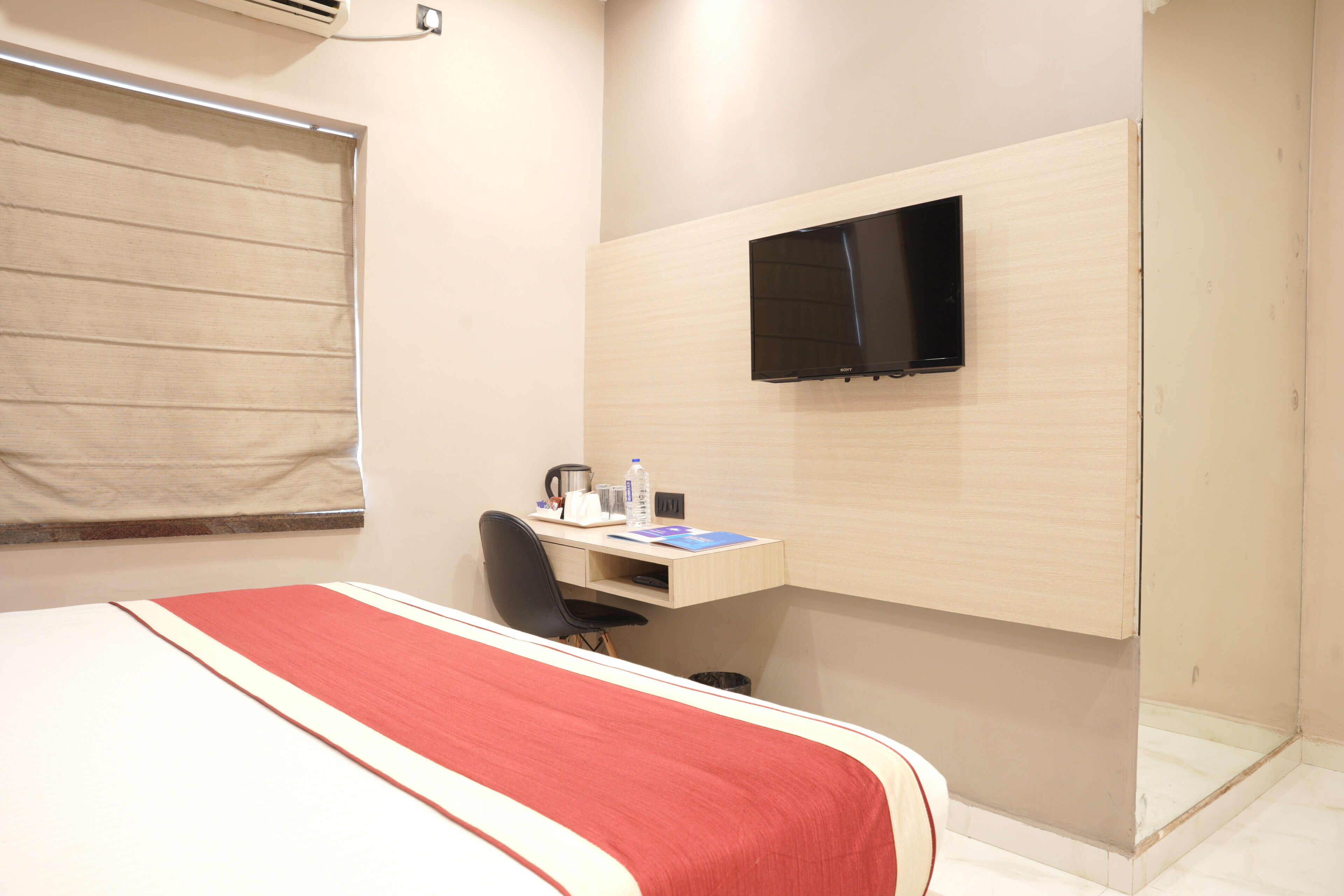 Quarto de hotel com cama, escrivaninha e cadeira, televisão de tela plana na parede e janela com persianas.
