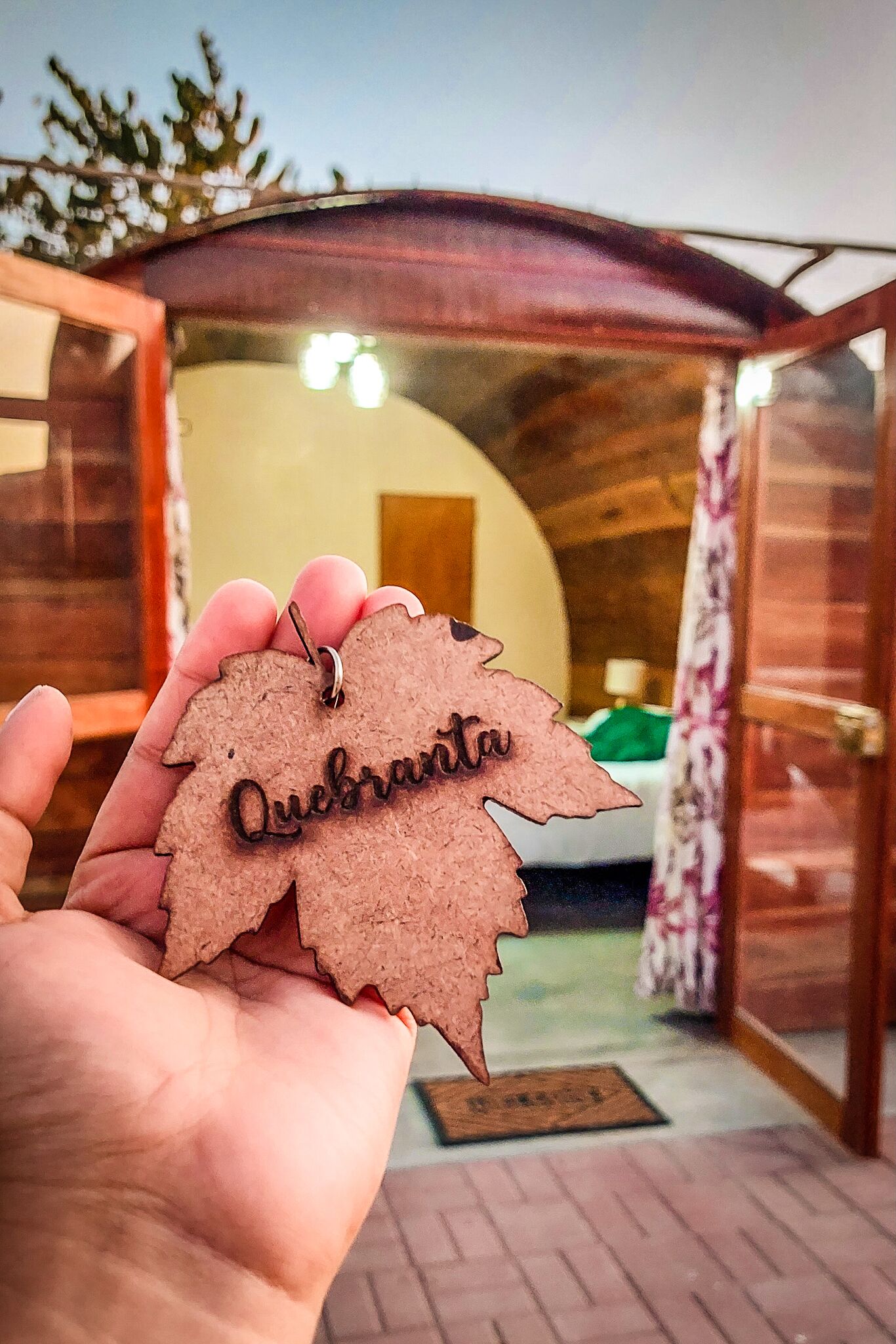Uma mão segura um chaveiro de madeira com o nome 'Quebranta' gravado. Ao fundo, uma estrutura de madeira com uma porta aberta mostra um quarto com uma cama e uma mesa de cabeceira.