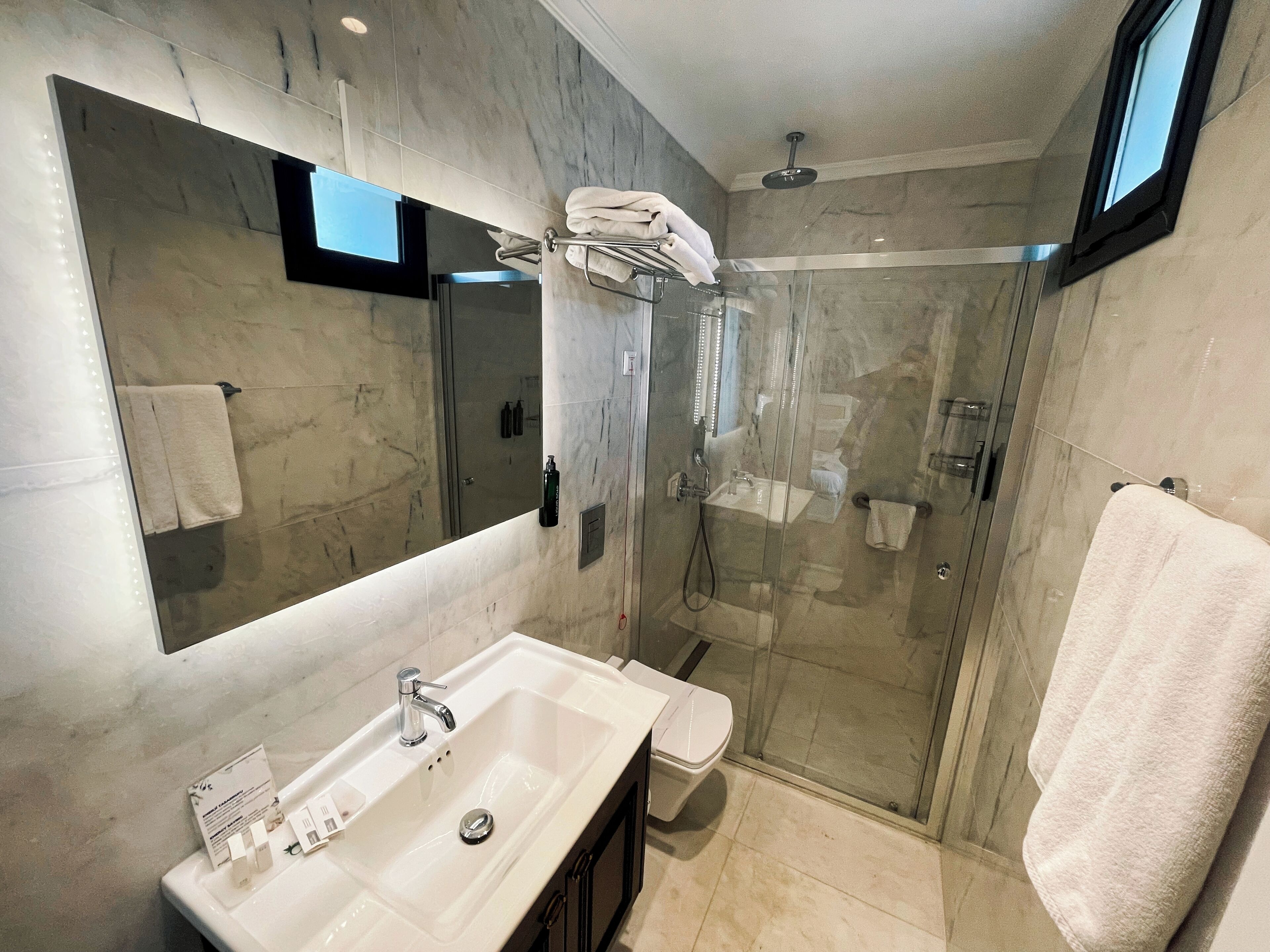 Casa de banho moderna com um espelho grande, uma sanita branca, uma cabine de duche com portas de vidro e um porta-toalhas.