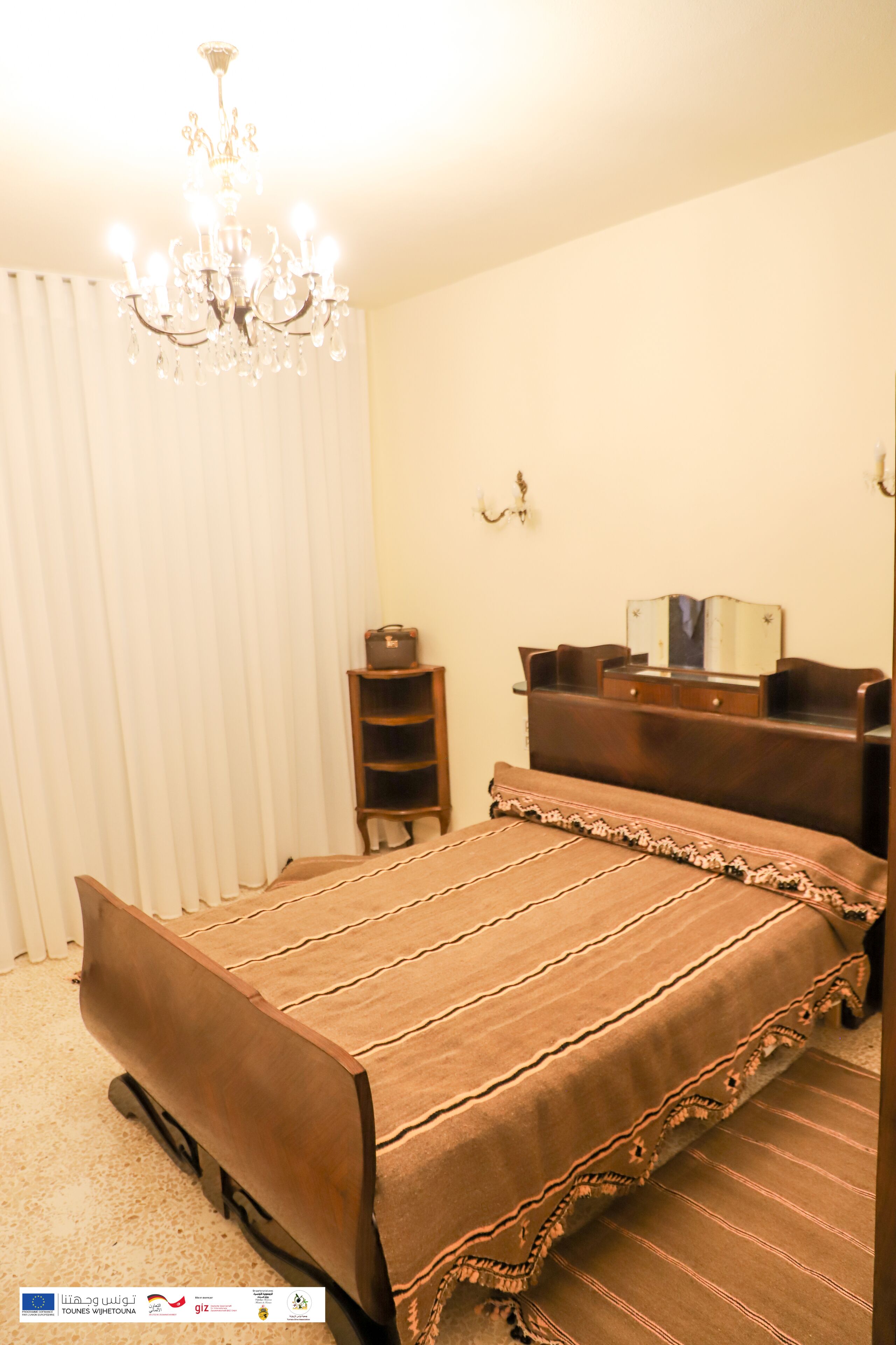 Quarto com cama de madeira, mesa de cabeceira, penteadeira, espelho e lustre.