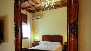 Quarto com um espelho grande e trabalhado, vigas de madeira no teto, cama com roupa de cama branca, mesa de cabeceira com abajur e criado-mudo pequeno.
