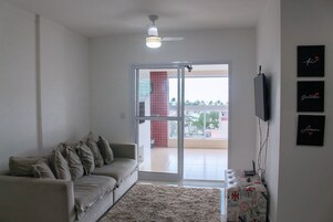 Uma sala de estar com sofá, TV de tela plana na parede e porta de vidro deslizante que dá para uma varanda.
