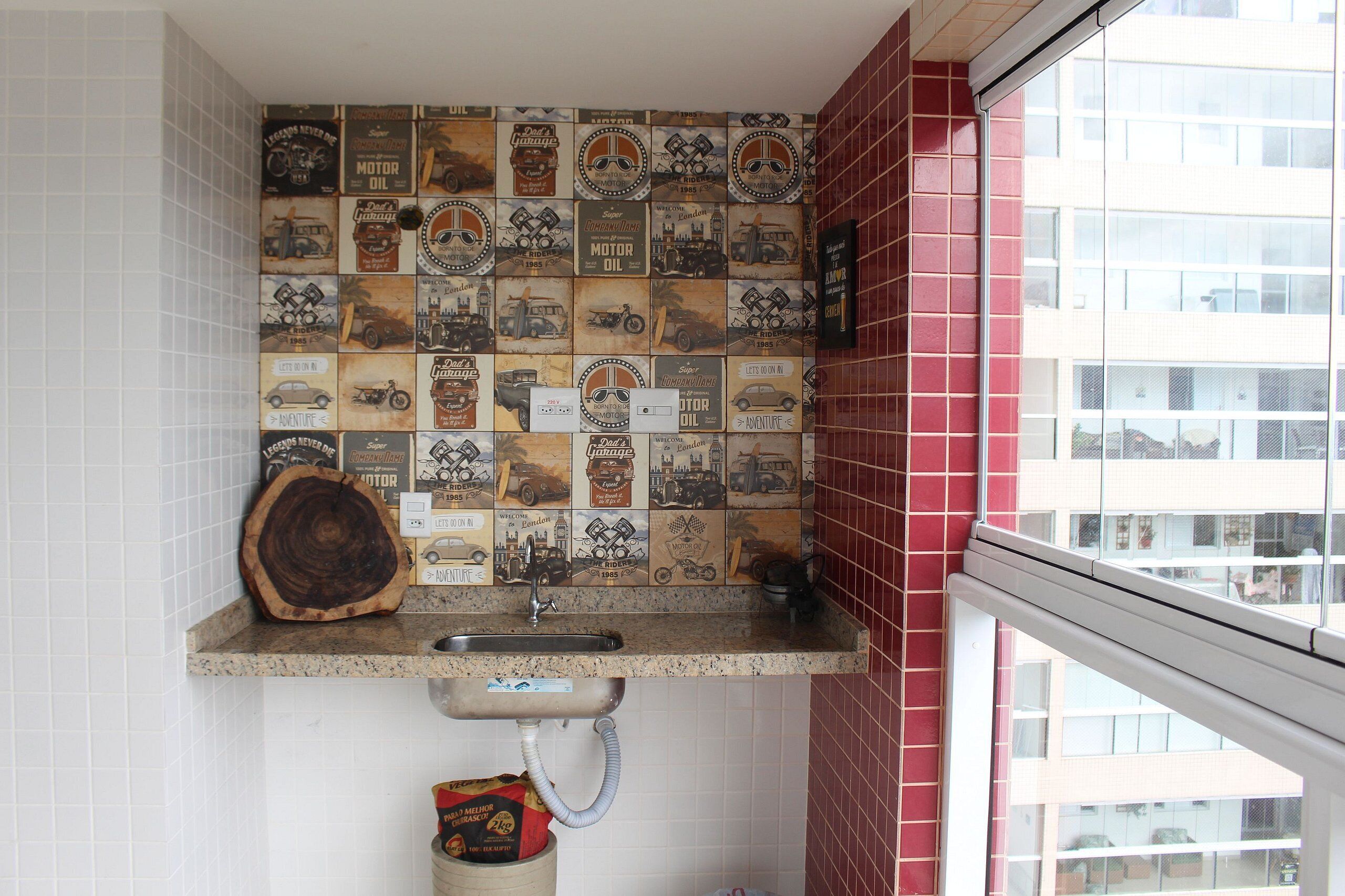 Cozinha com painel de azulejos com desenhos de automóveis antigos, uma pia de aço inoxidável e uma janela com vista para um edifício.