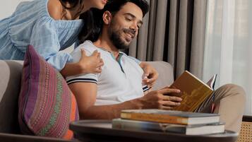 Um casal sentado em um sofá com um livro, uma mesinha com livros e uma cadeira de vime.