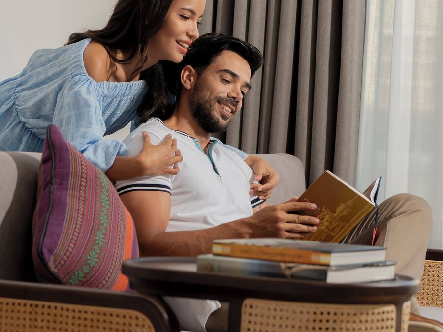 Um casal sentado em um sofá com um livro, uma mesinha com livros e uma cadeira de vime.