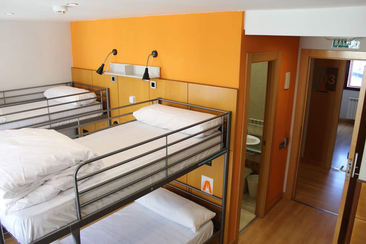 Quarto tipo dormitório com beliches, uma pequena estante com abajur e uma porta que dá para um banheiro.