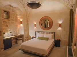 Quarto com parede de pedra, espelho redondo, cama com almofadas, mesa de cabeceira e luminária pendente.
