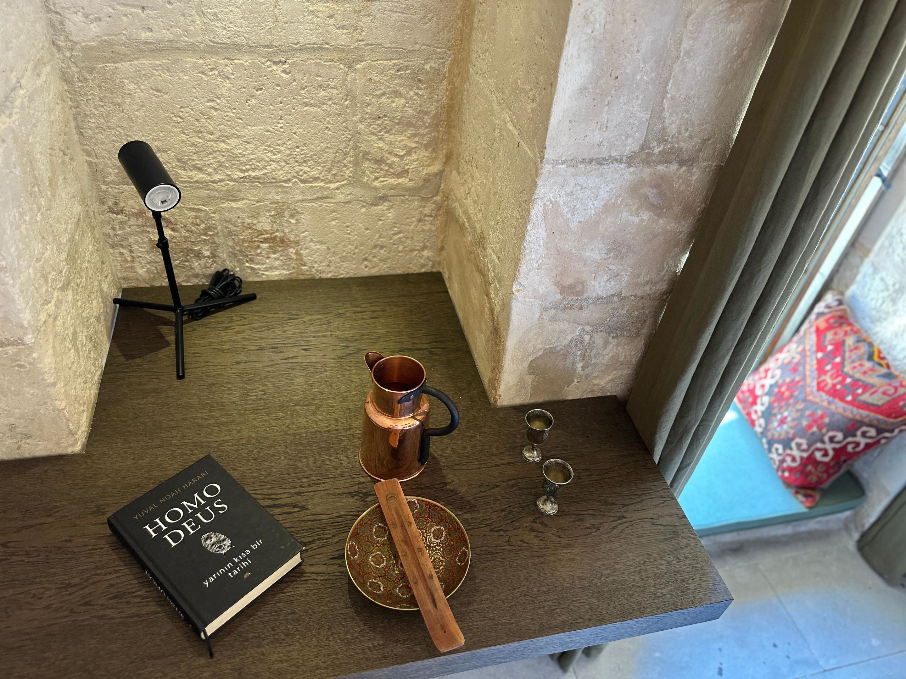 Uma pequena mesa de madeira com um livro intitulado 'Homo Deus' de Yuval Harari, uma taça de cobre, uma colher de madeira e uma pequena taça de metal.