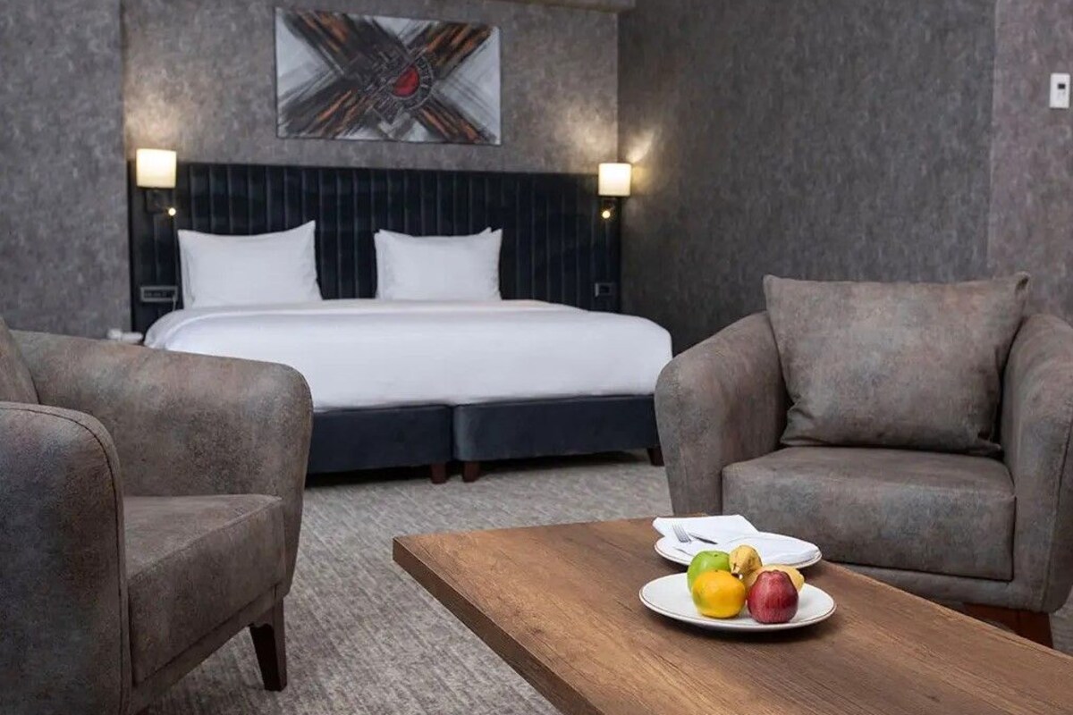 Quarto de hotel com cama, duas poltronas, mesa de centro de madeira com frutas e um quadro na parede.