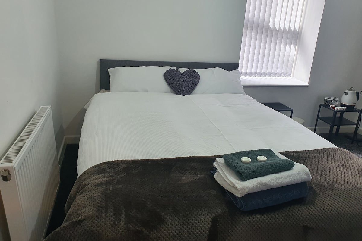 Cama bem arrumada com roupa de cama branca, um travesseiro em formato de coração e um cobertor marrom.